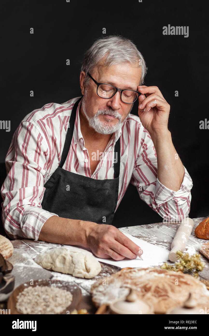 Old Baker writing down old-time recipe in bakery notebook surrounded by ...