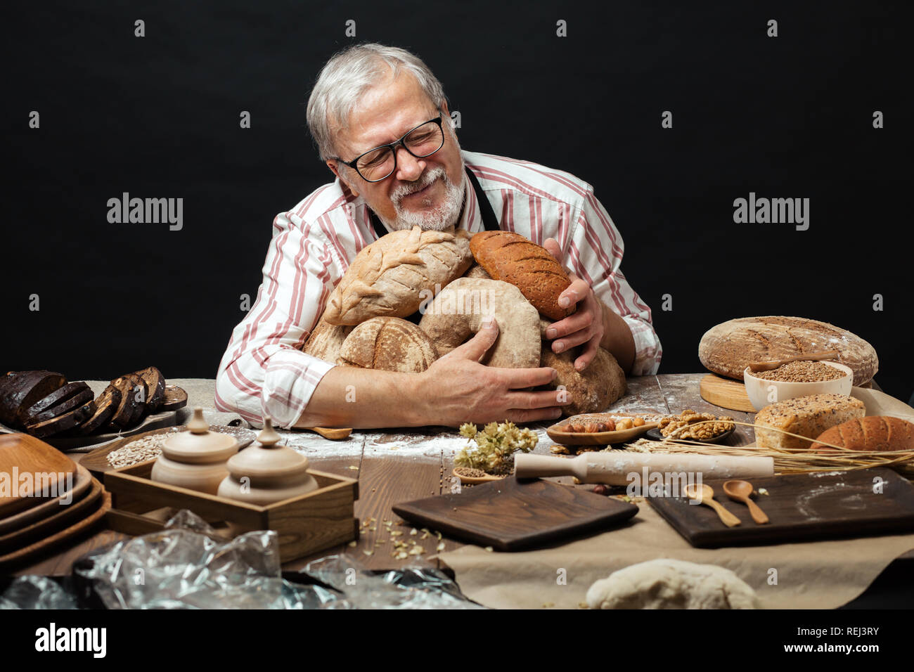Happy old baker looking at camera and smiling while hugging loaves of ...