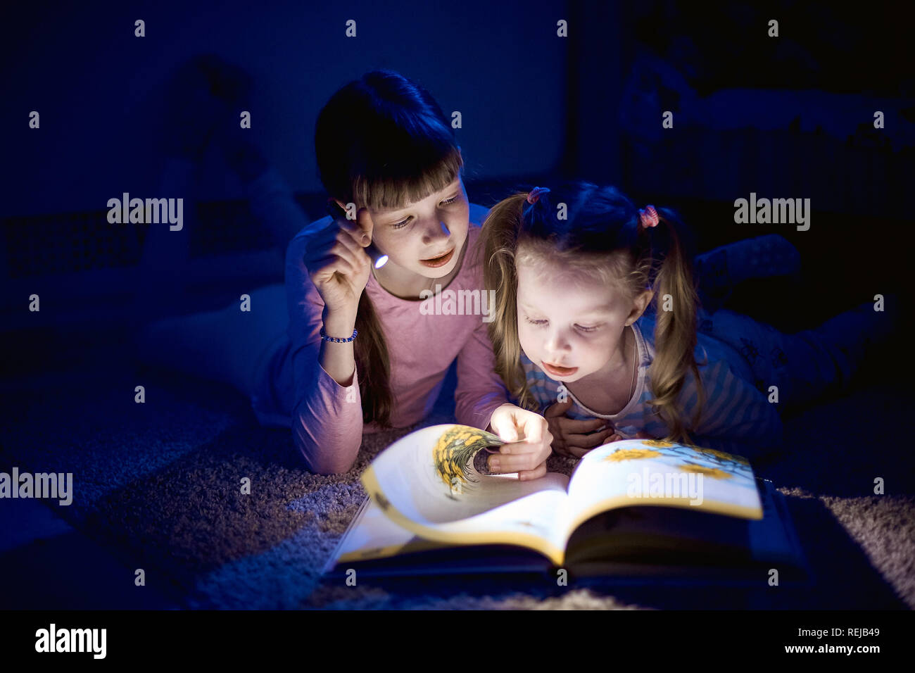 two sisters reading a book with a flashlight in a dark room at night ...