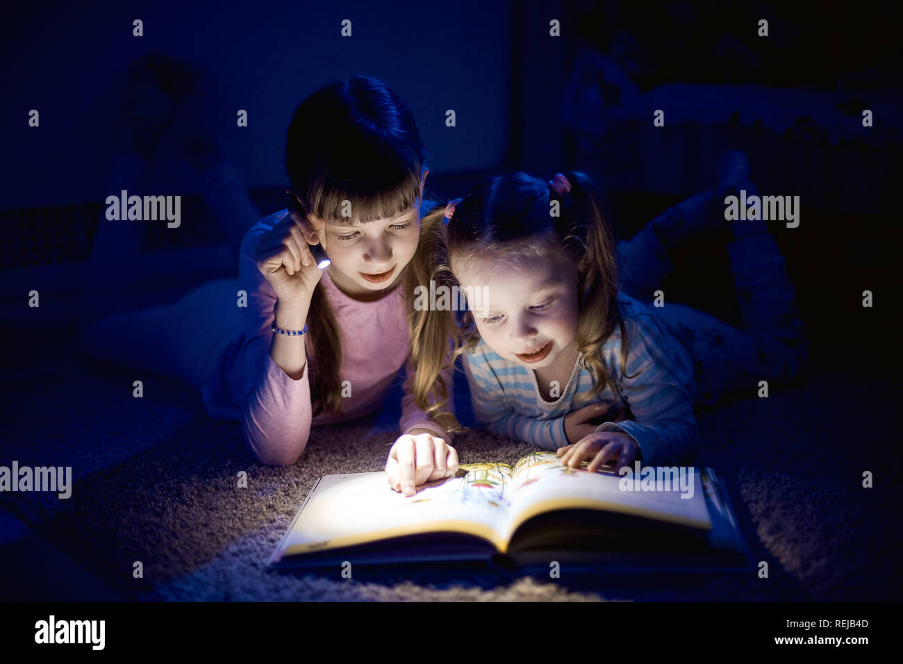 two sisters reading a book with a flashlight in a dark room at night ...