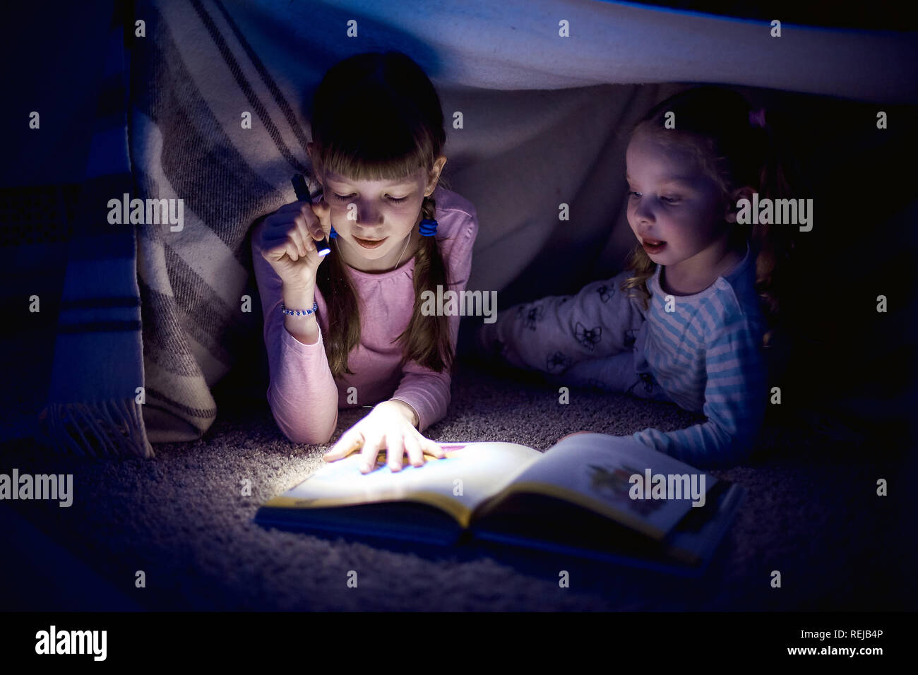 two sisters reading a book with a flashlight in a dark room at night ...