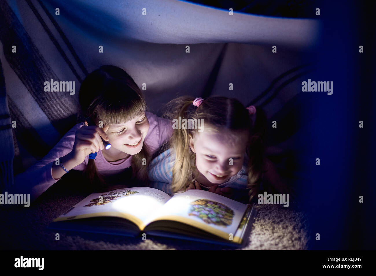 two sisters reading a book with a flashlight in a dark room at night ...