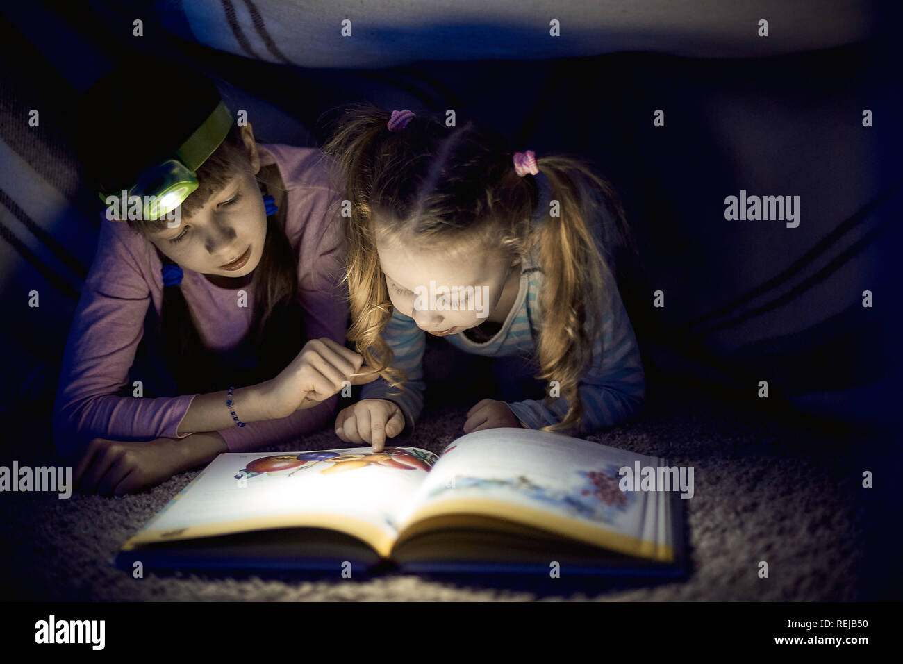 two sisters reading a book with a flashlight in a dark room at night ...