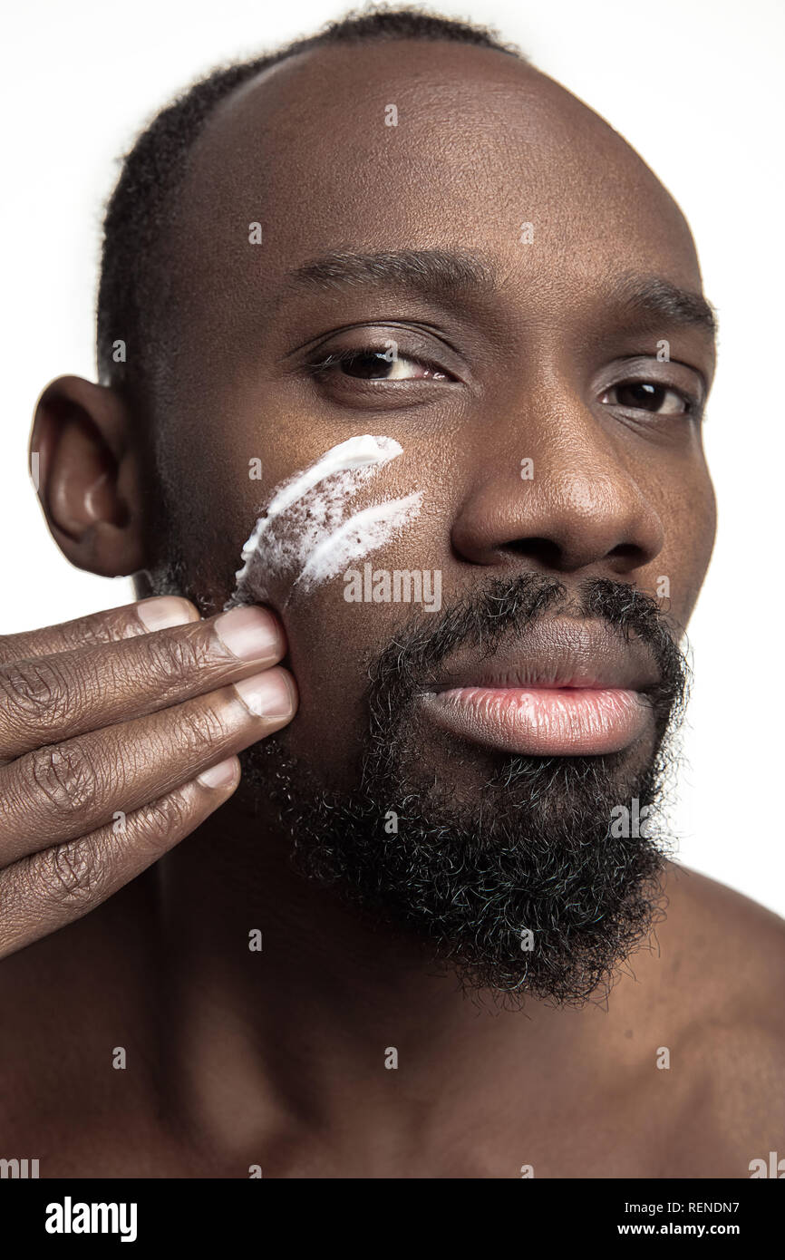 Young african-american guy applying face cream under his eyes on white ...