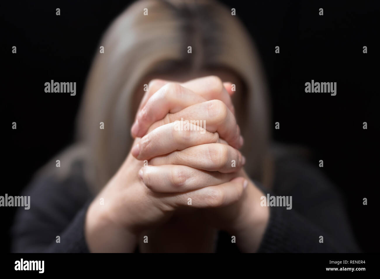 Woman praying with her hands clasped in front of her face, in dark room ...