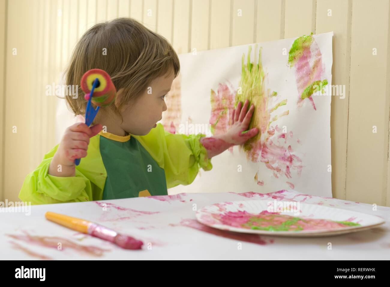 A two year old girl in a plastic smock painting in a playroom with a ...