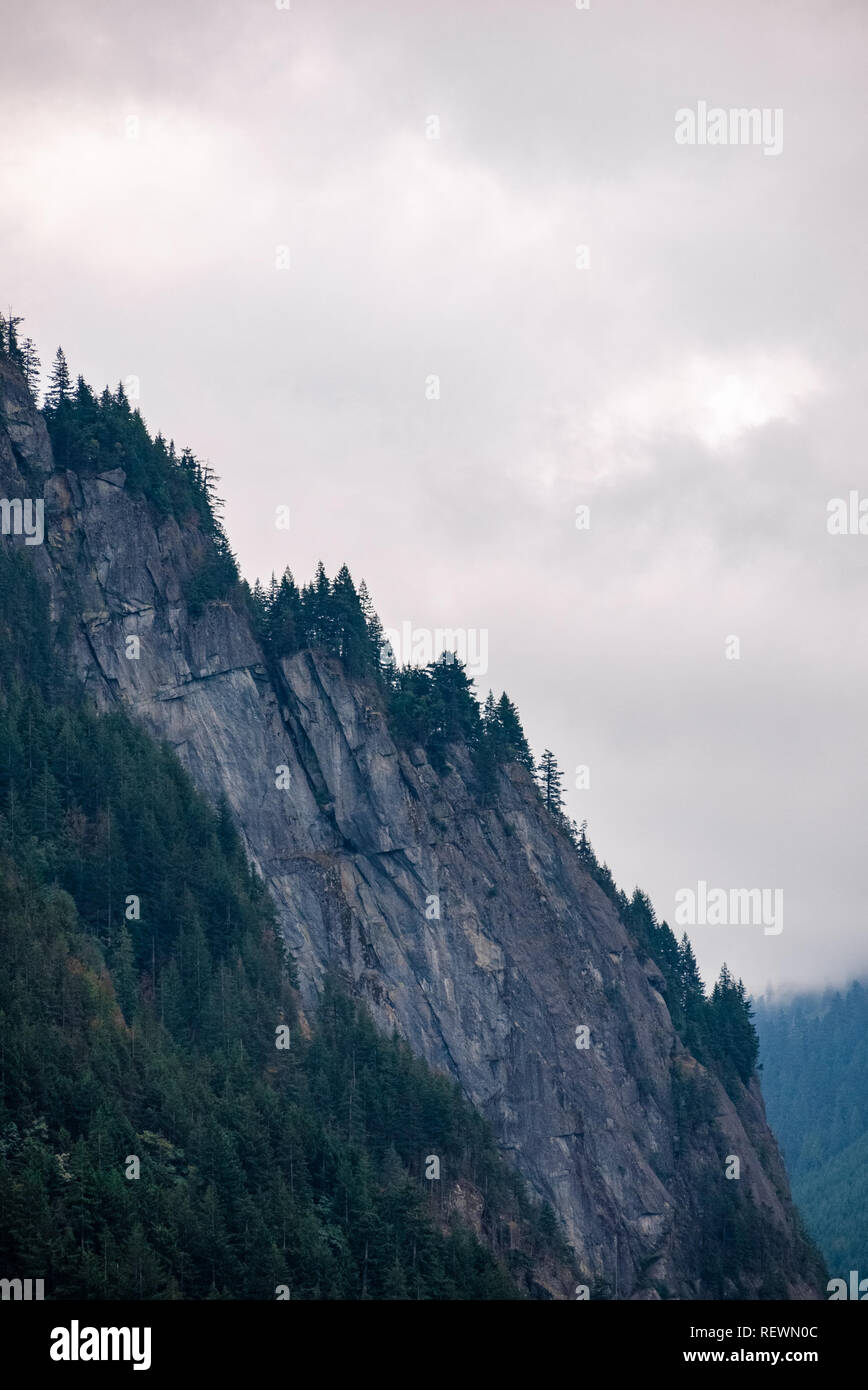 Steep rocky cliff of mountain surrounded by trees on overcast sky ...