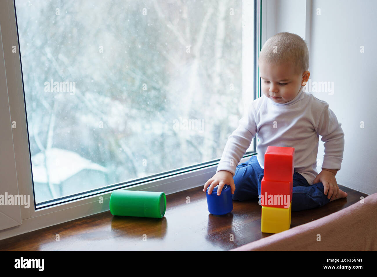 Little boy sitting on window sill. He builds tower of cubes. Winter day ...