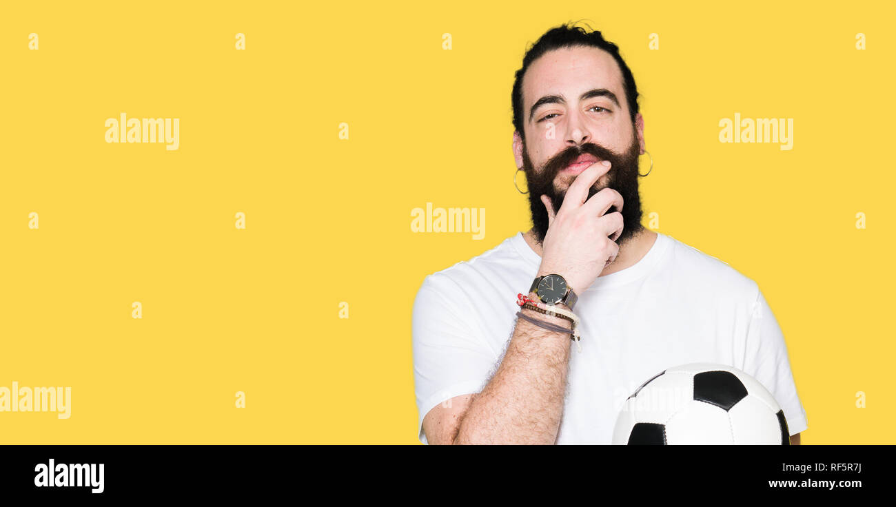 Young man with long hair and bear holding soccer football ball serious ...