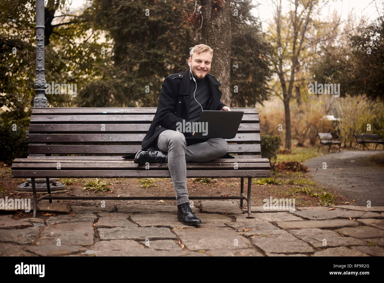 one young smiling and happy man, sitting on bench in public park, using ...