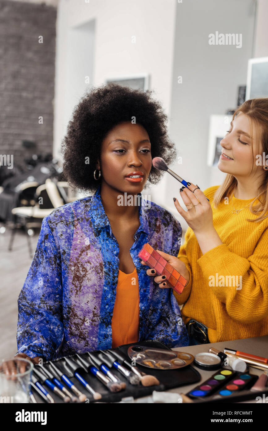 Fair-haired positive stylist doing makeup to the dark-skinned model ...