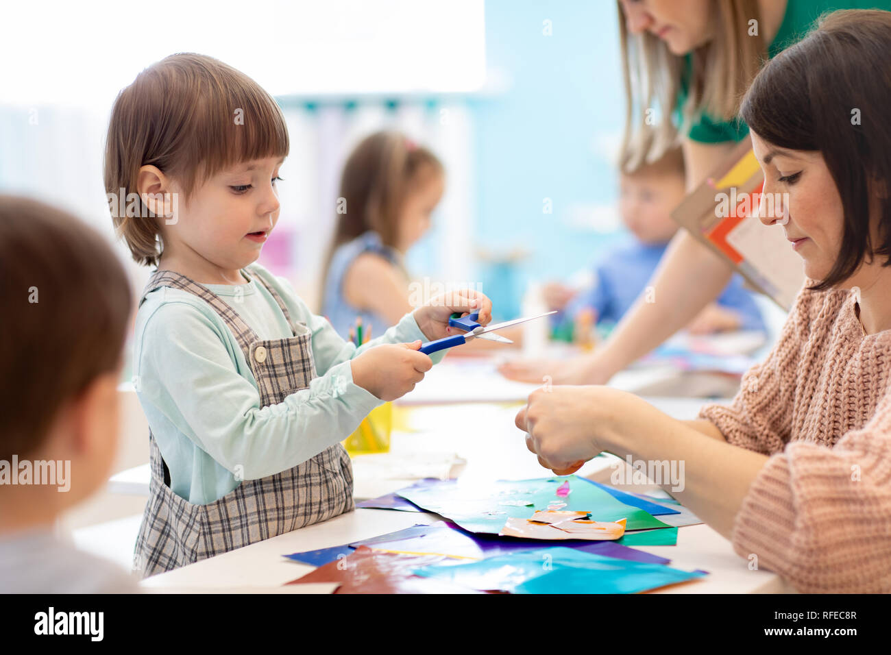 Child with scissors in hands cutting paper with teachers in class room ...