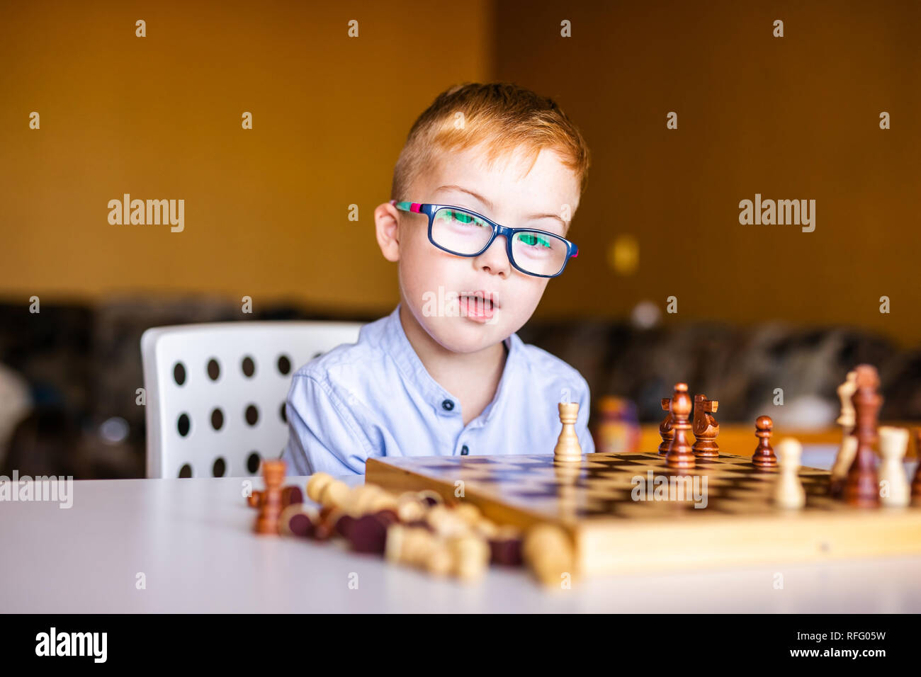Ginger boy with down syndrome with big glasses playing chess at home ...