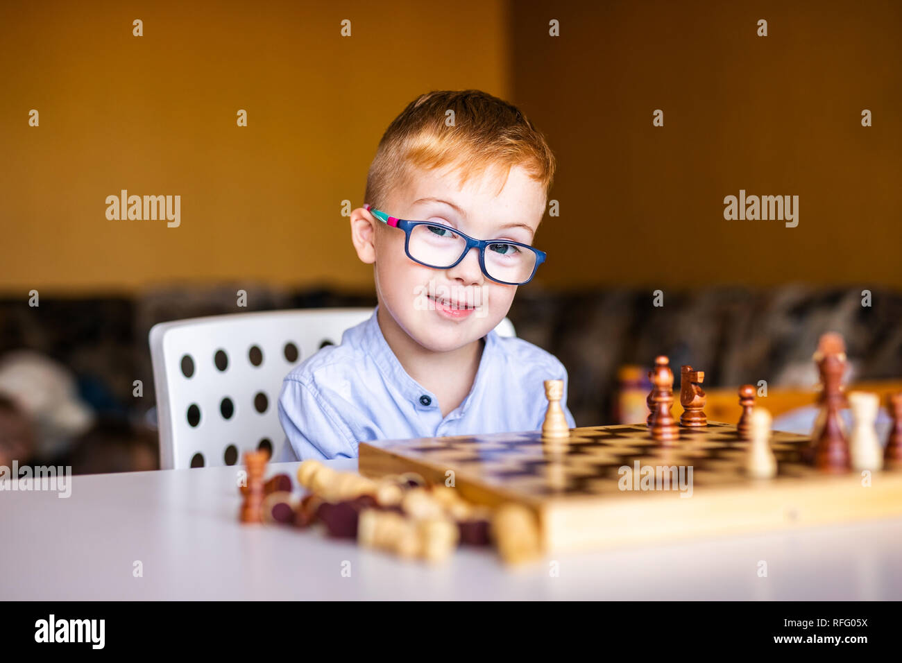 Ginger boy with down syndrome with big glasses playing chess at home ...