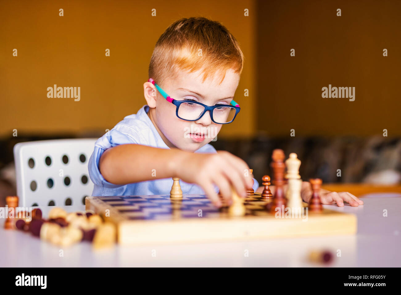 Ginger boy with down syndrome with big glasses playing chess at home ...