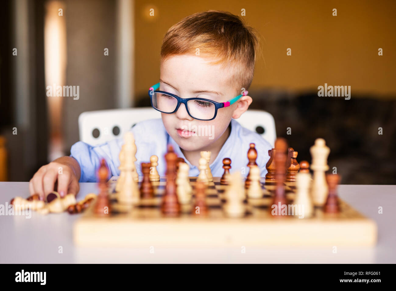 Ginger boy with down syndrome with big glasses playing chess at home ...