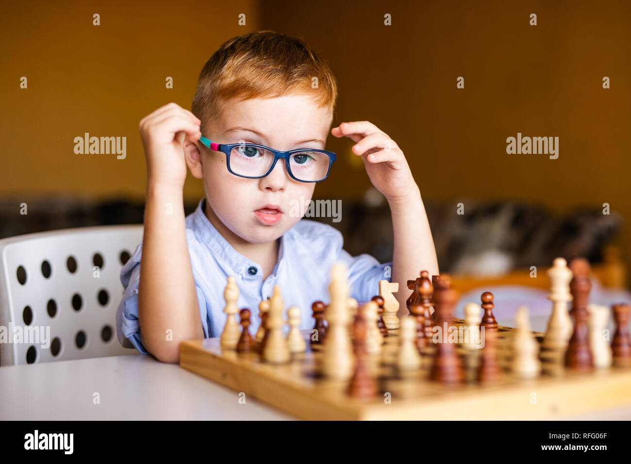 Ginger boy with down syndrome with big glasses playing chess at home ...