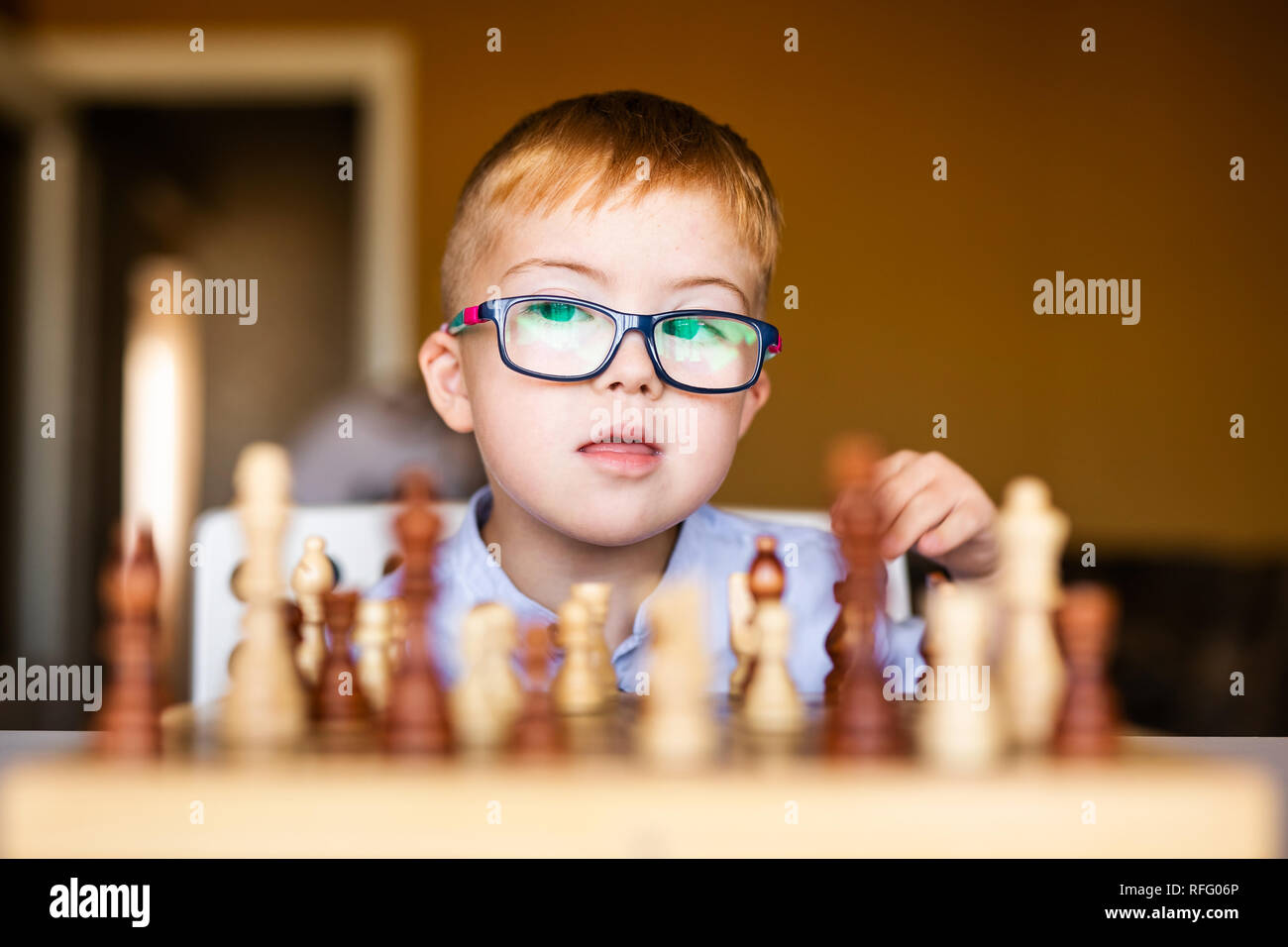 Ginger boy with down syndrome with big glasses playing chess at home ...