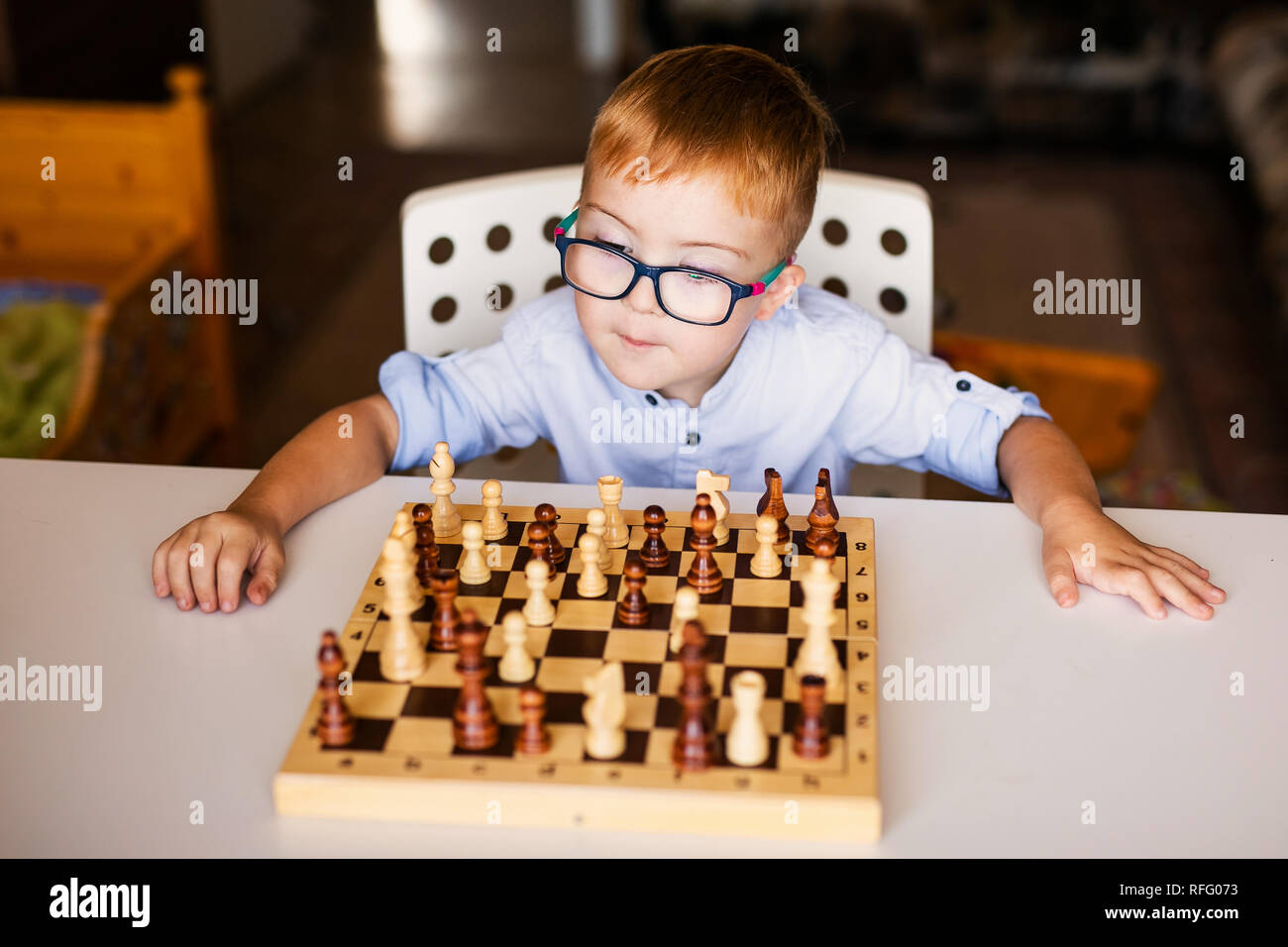 Ginger boy with down syndrome with big glasses playing chess at home ...