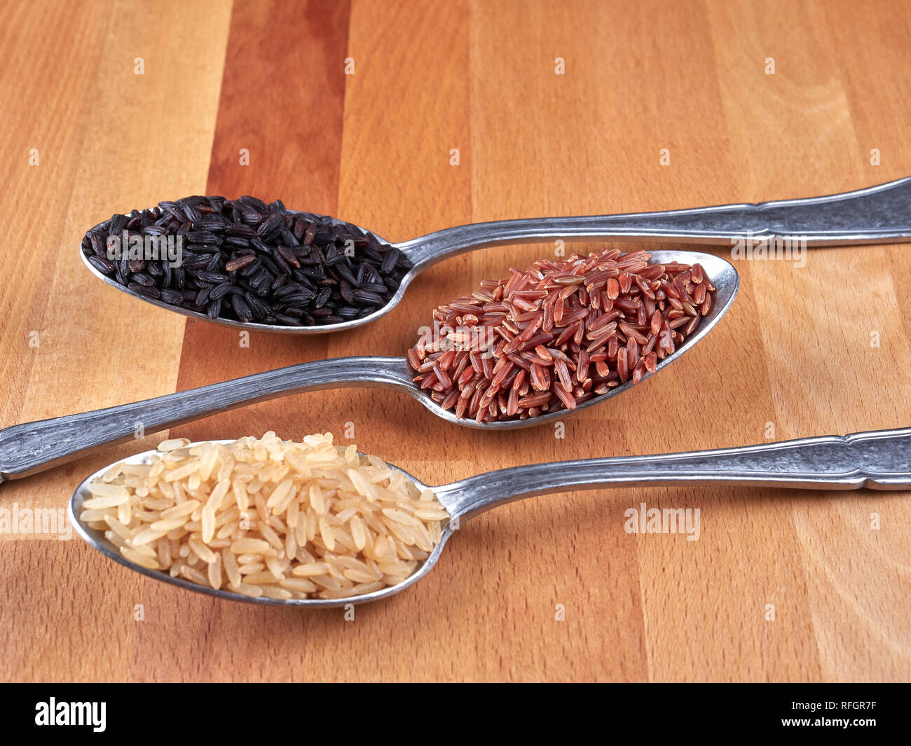 Uncooked black, red and brown rice on silver spoons, on a rustic wooden ...