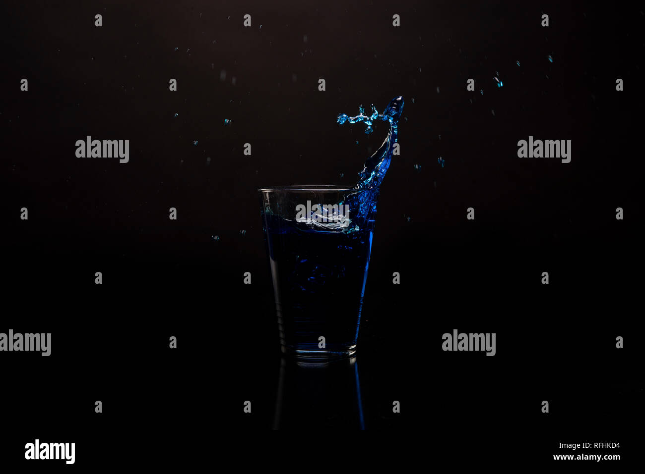Glass blue drink splashing out. Shot against black reflective surface ...