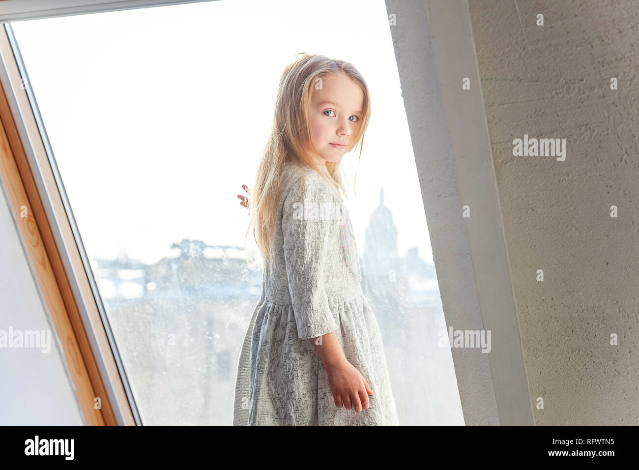 Little cute sweet smiling girl in white dress standing on the window ...