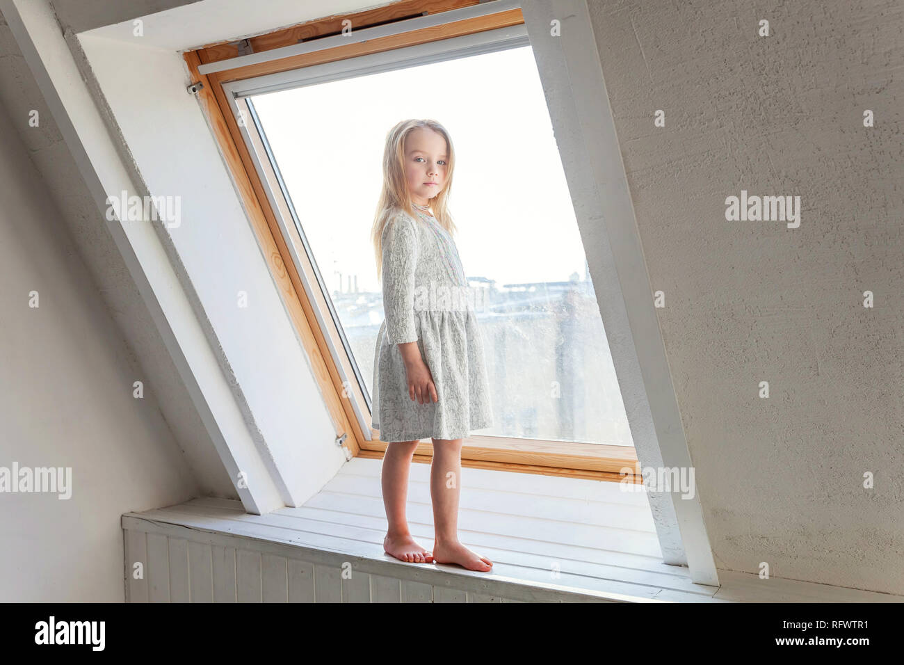 Little cute sweet smiling girl in white dress standing on the window ...
