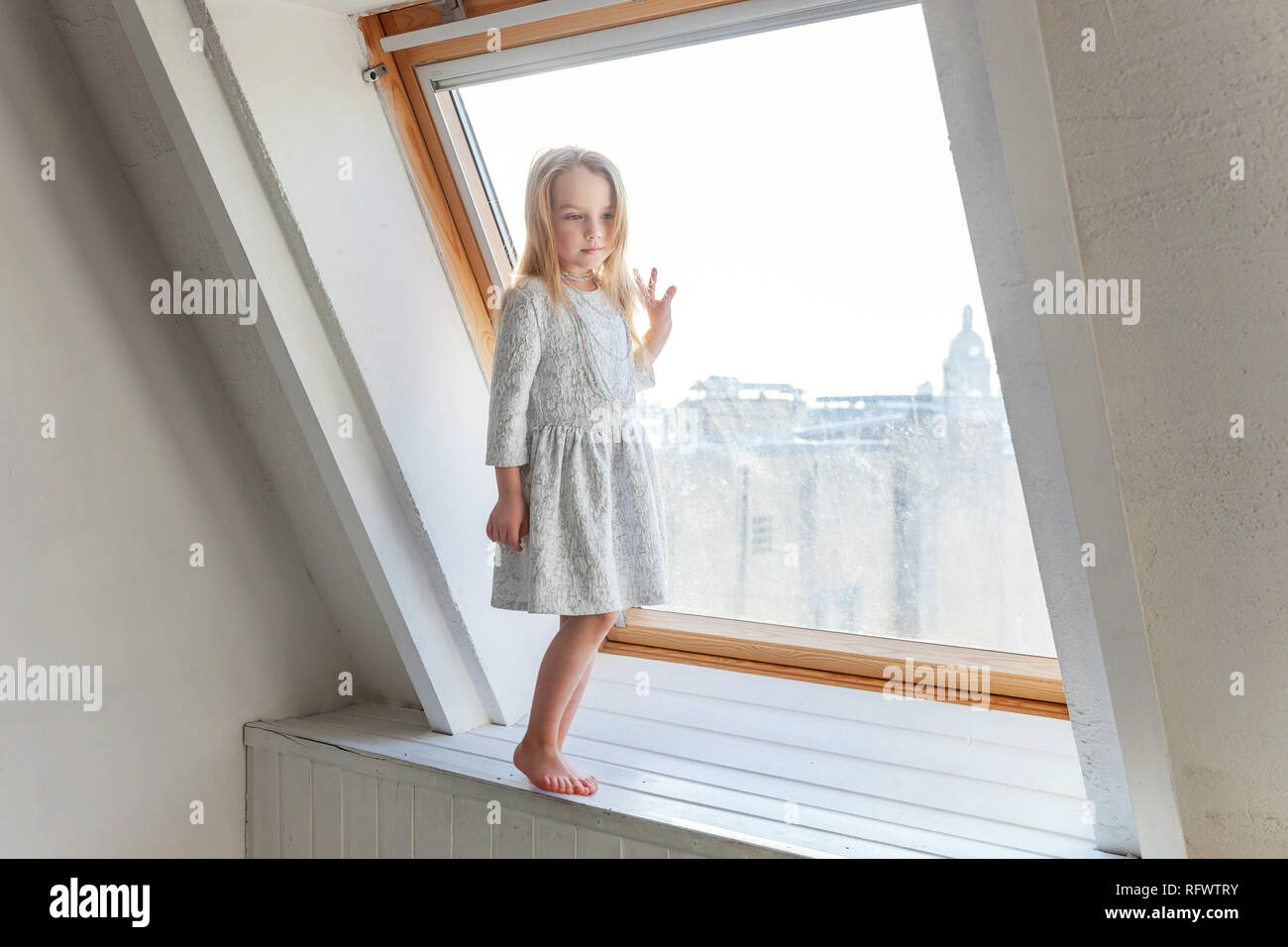 Little cute sweet smiling girl in white dress standing on the window ...