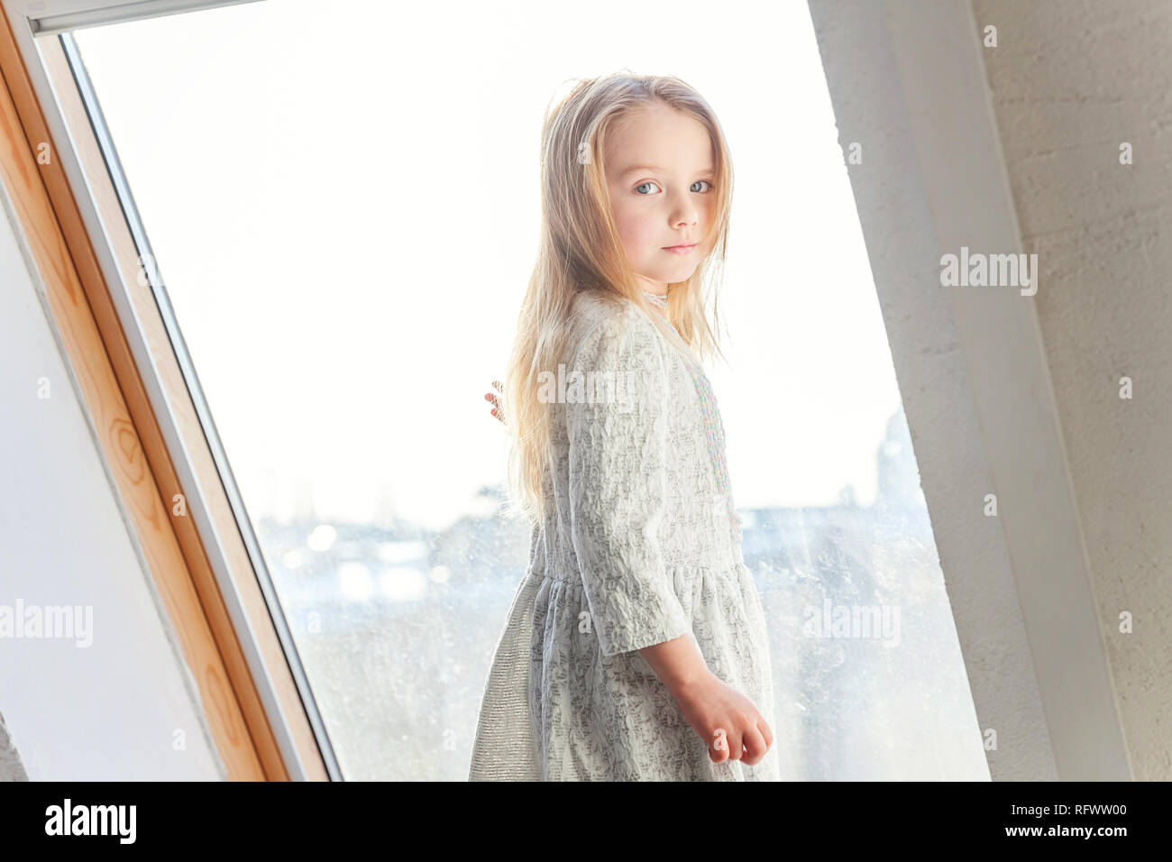Little cute sweet smiling girl in white dress standing on the window ...