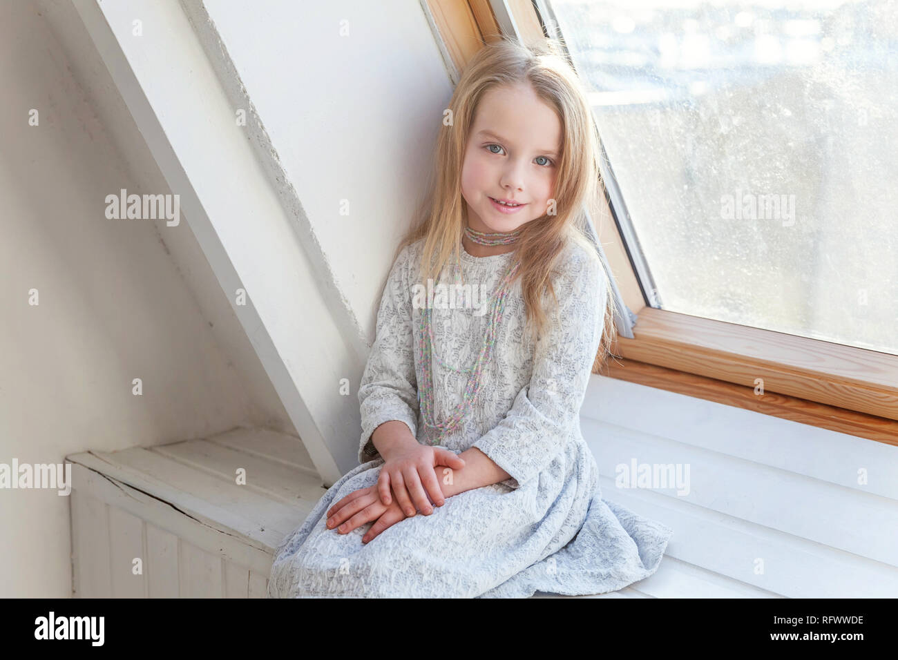 Little cute sweet smiling girl in white dress sitting on the window ...