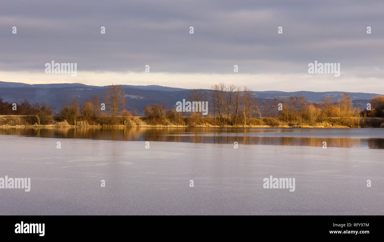 Frozen lake, sunlit, golden trees without leaves and horizon mountains ...