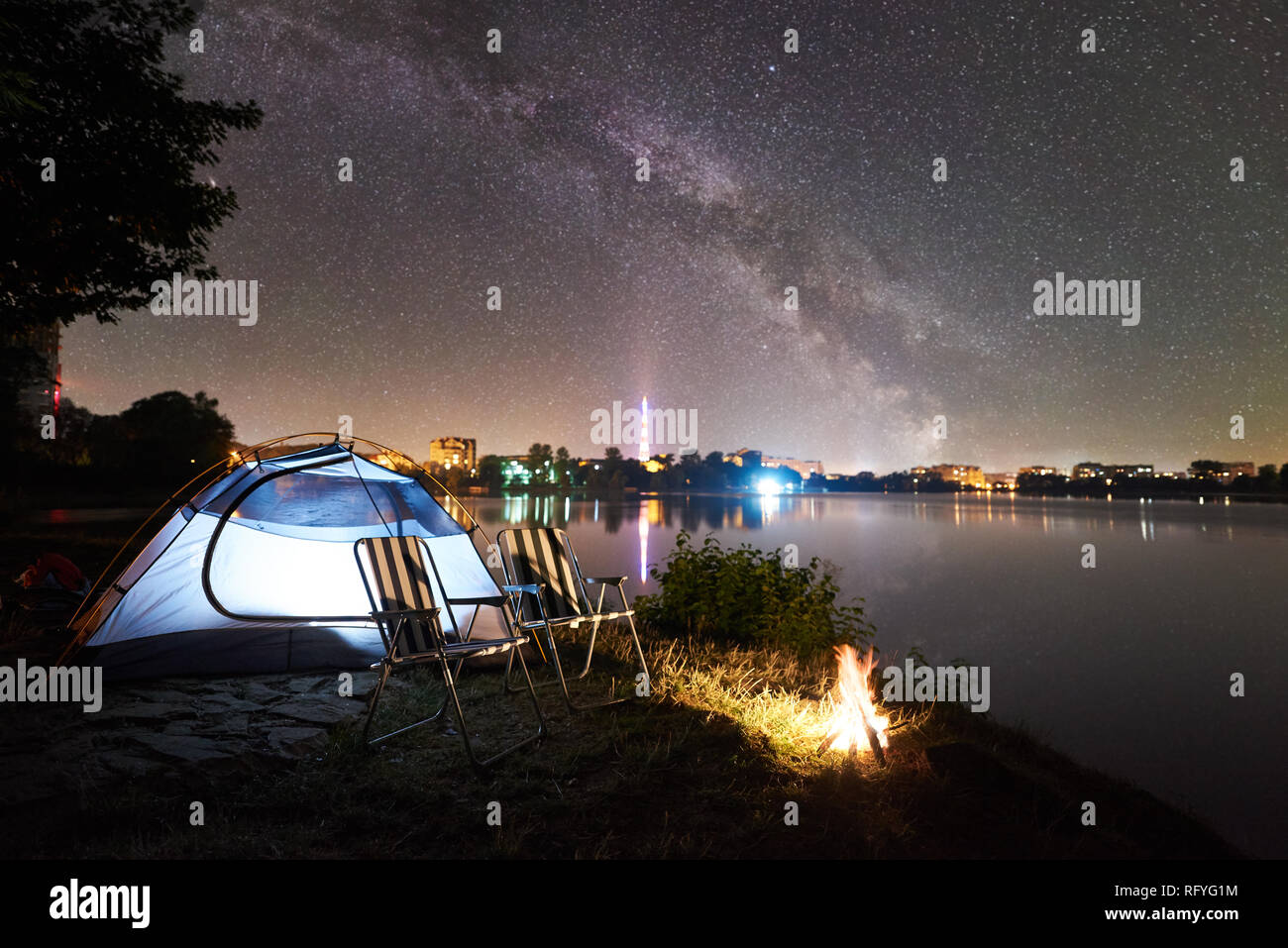 Night camping. Illuminated tent, two chairs and campfire on lake shore ...