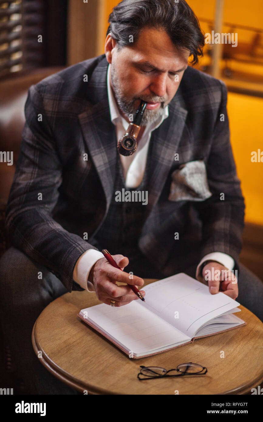 Mature businessman smoking tobacco pipe and making notes, working at ...
