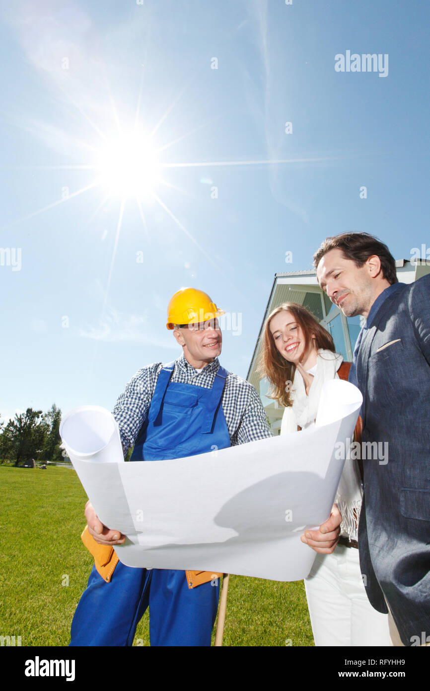 Worker shows house design plans to a young couple at construction site ...