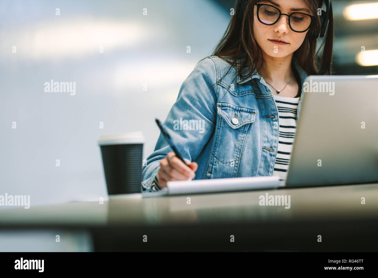 University student writing in a book while sitting at desk with laptop ...
