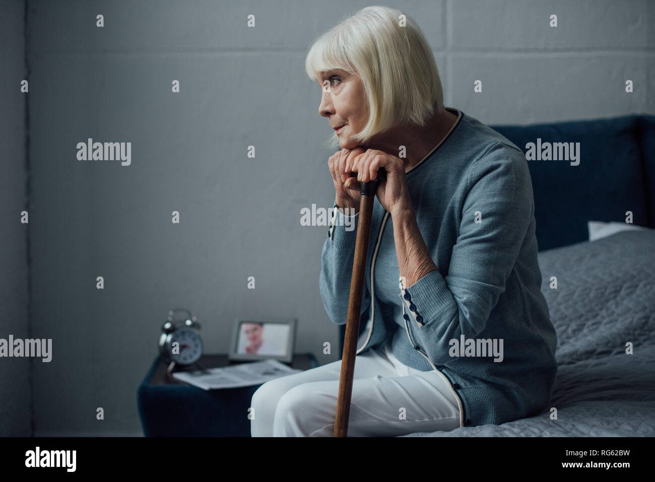 lonely senior woman with hands on walking stick sitting on bed at home ...