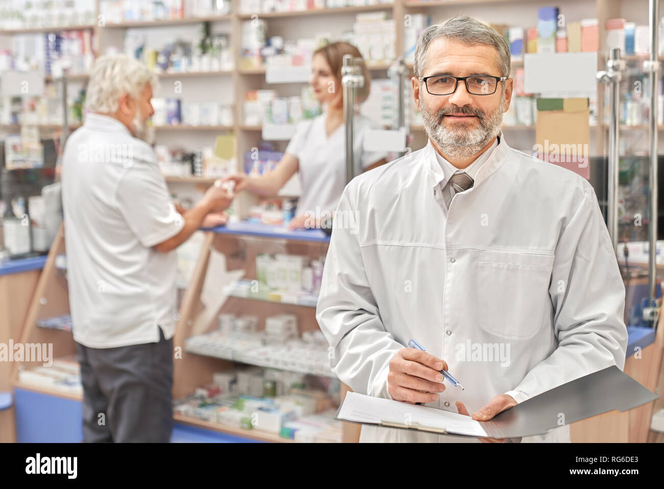 Happy pharmacist posing in pharmacy with folder in hands. Bearded man ...