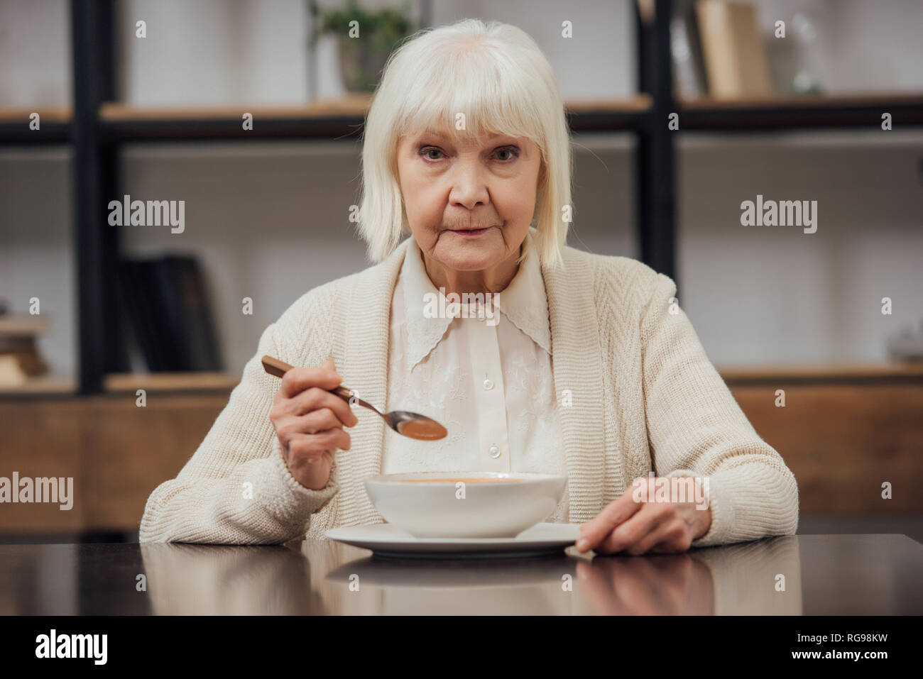 sad senior woman sitting at table, looking at camera and eating at home ...