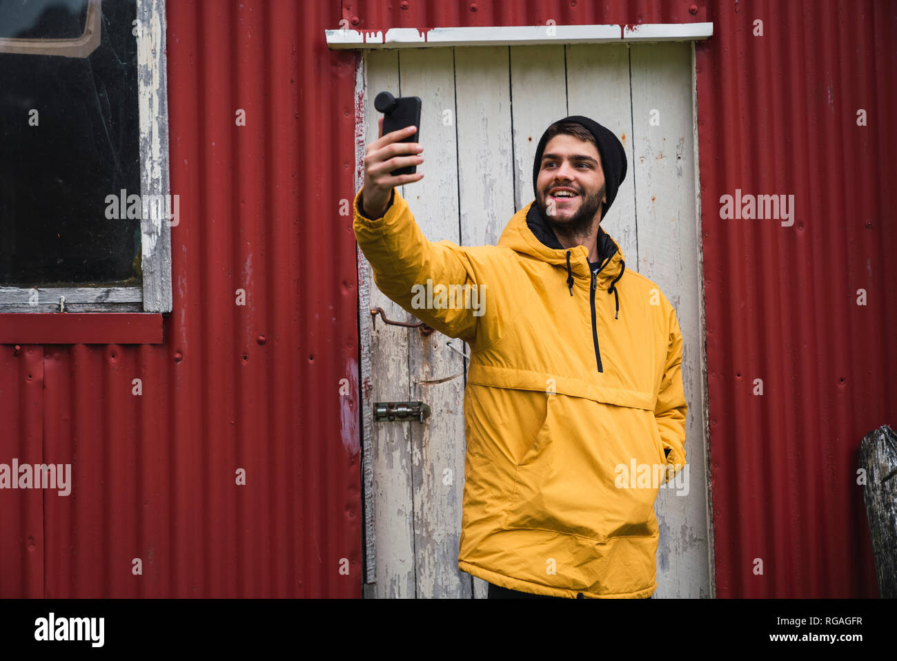 Young man standing in front of corrugated iron shack, taking selfie ...