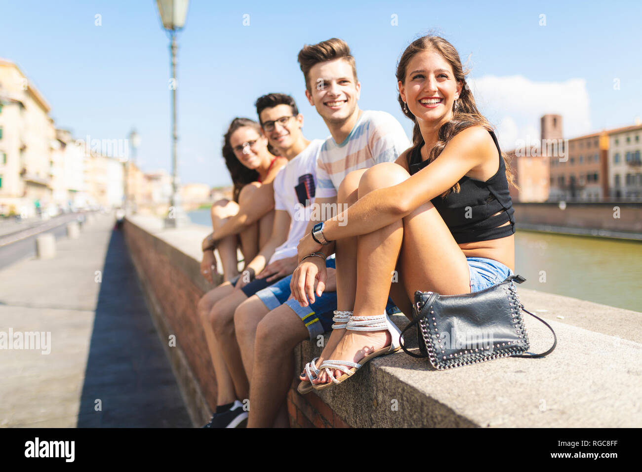 Italy, Pisa, group of four happy friends sitting on a wall along Arno ...