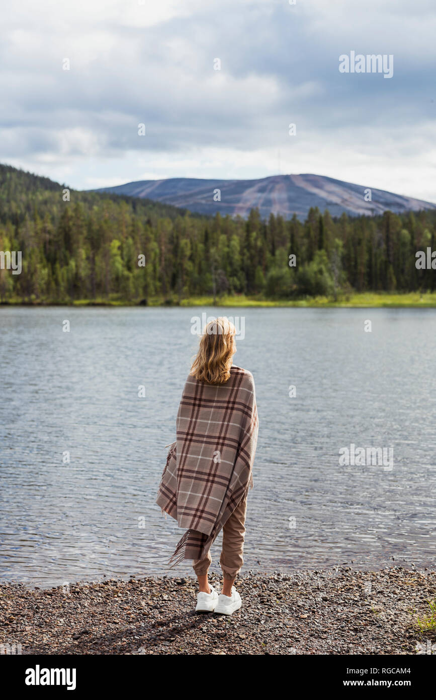 Finland, Lapland, woman wrapped in a blanket standing at the lakeside ...