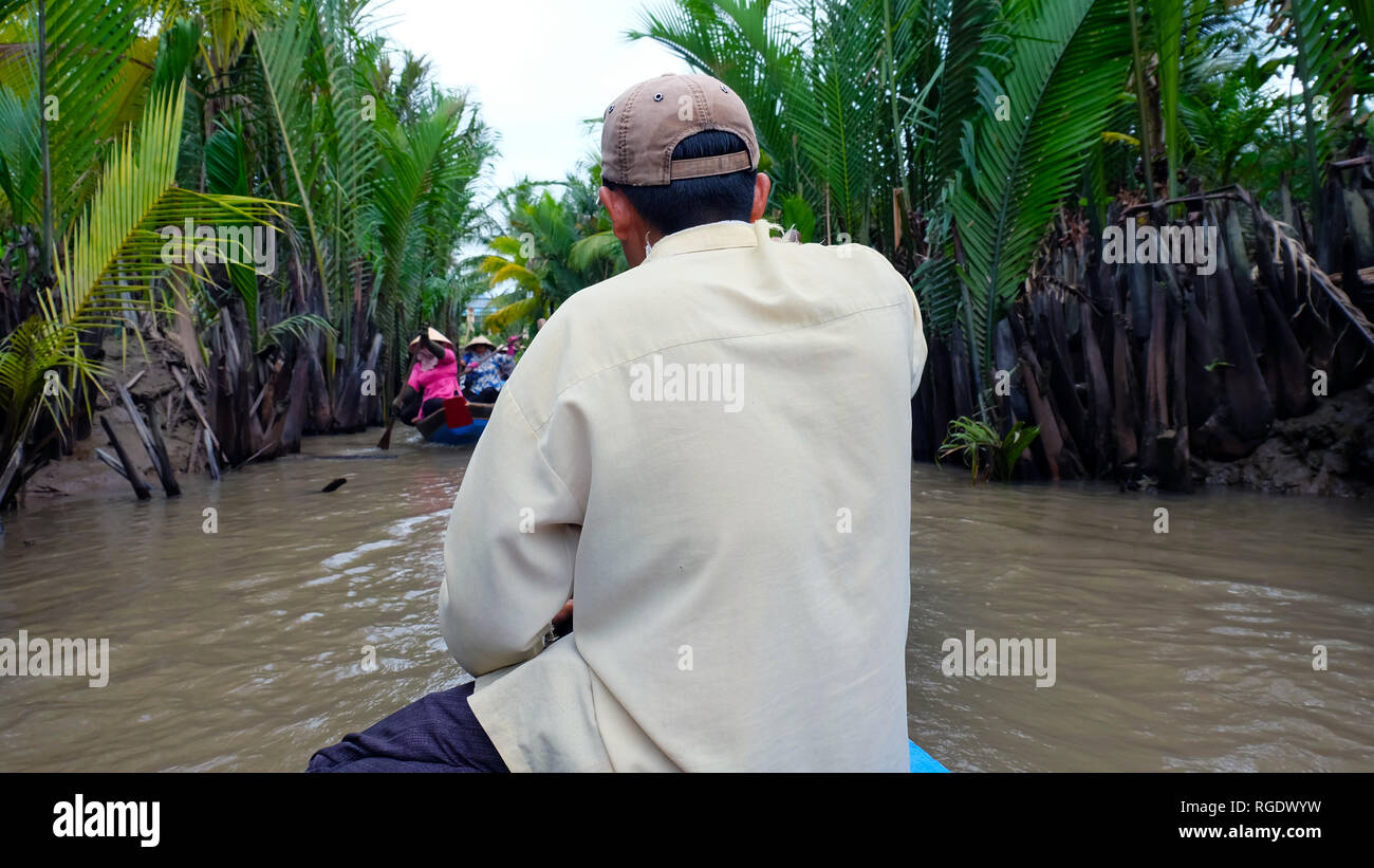 Back of a man, sitting at the front of a boat, rowing along a muddy ...