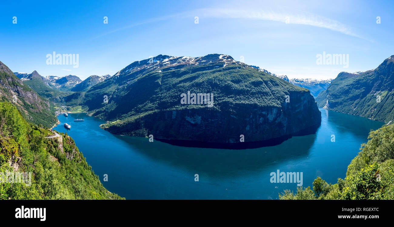A view of a green, deep fjord surrounded by mountains under a blue sky ...