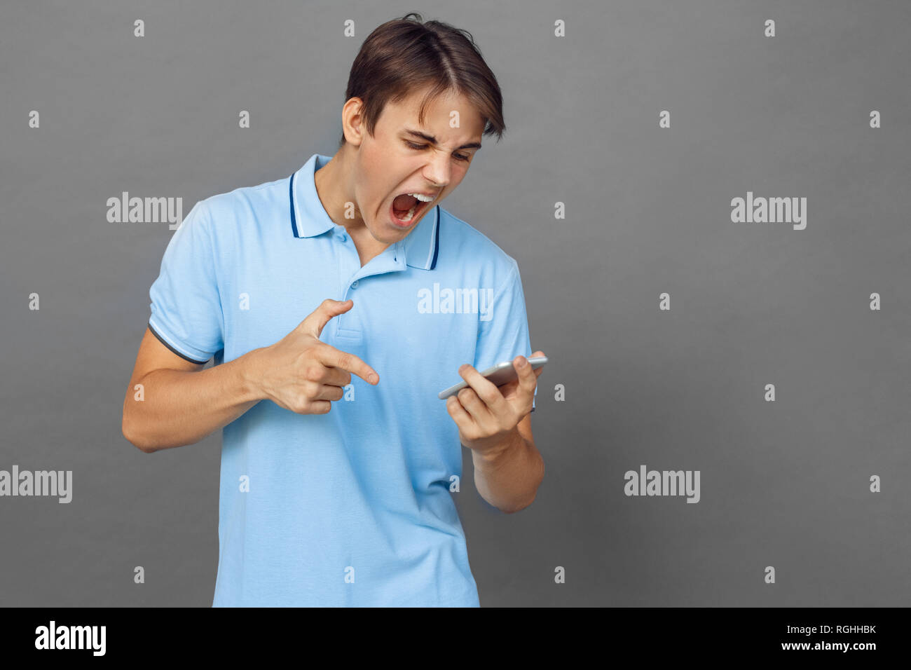 Young man standing isolated on grey wall shouting at smartphone angry ...