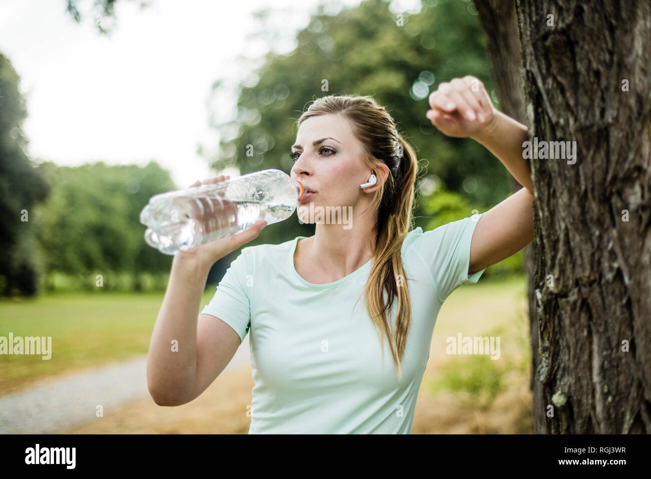 Sportive young woman leaning against a tree in a park drinking from ...