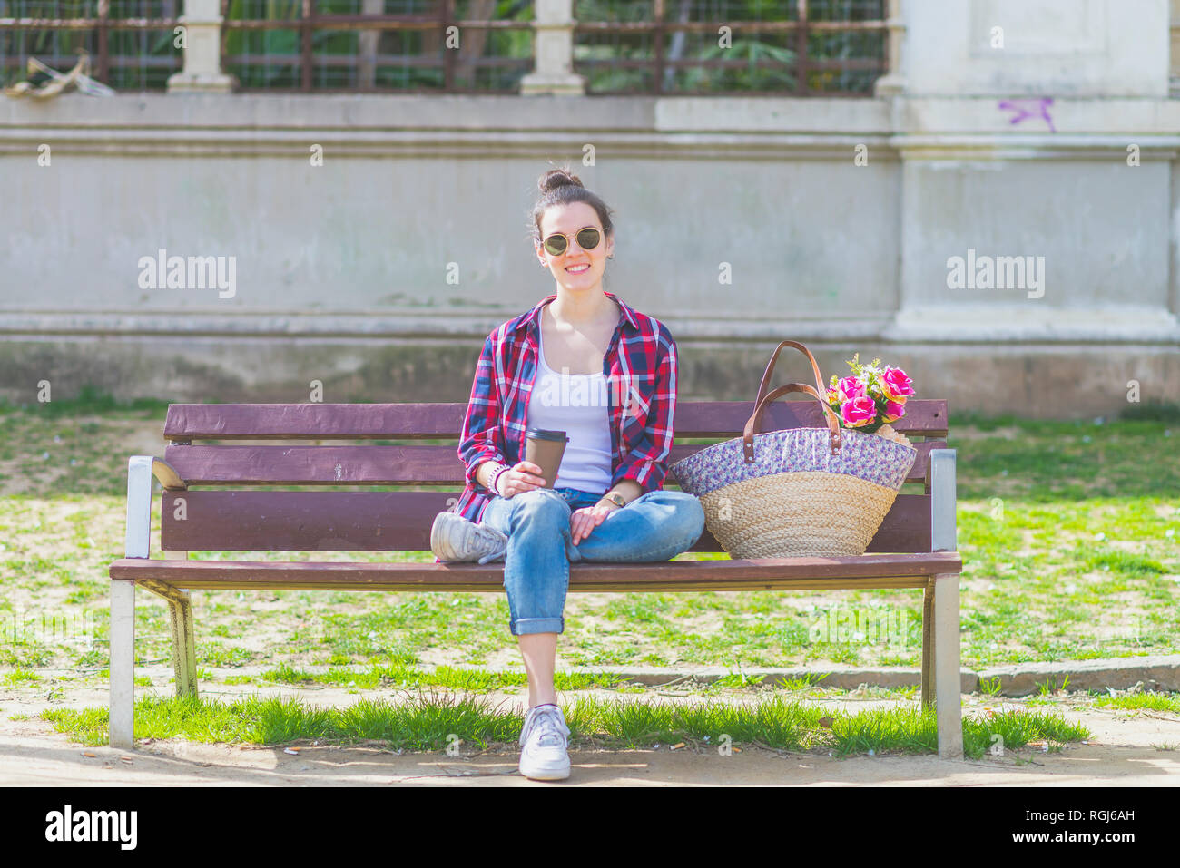 Front view of a young hipster Woman sitting on a park bench relaxing in ...