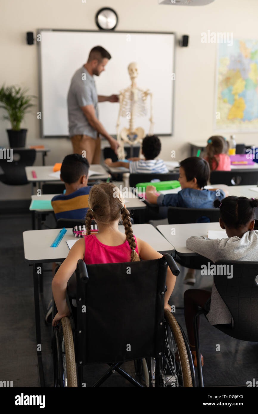 Rear view of disable schoolgirl with classmate studying in classroom ...