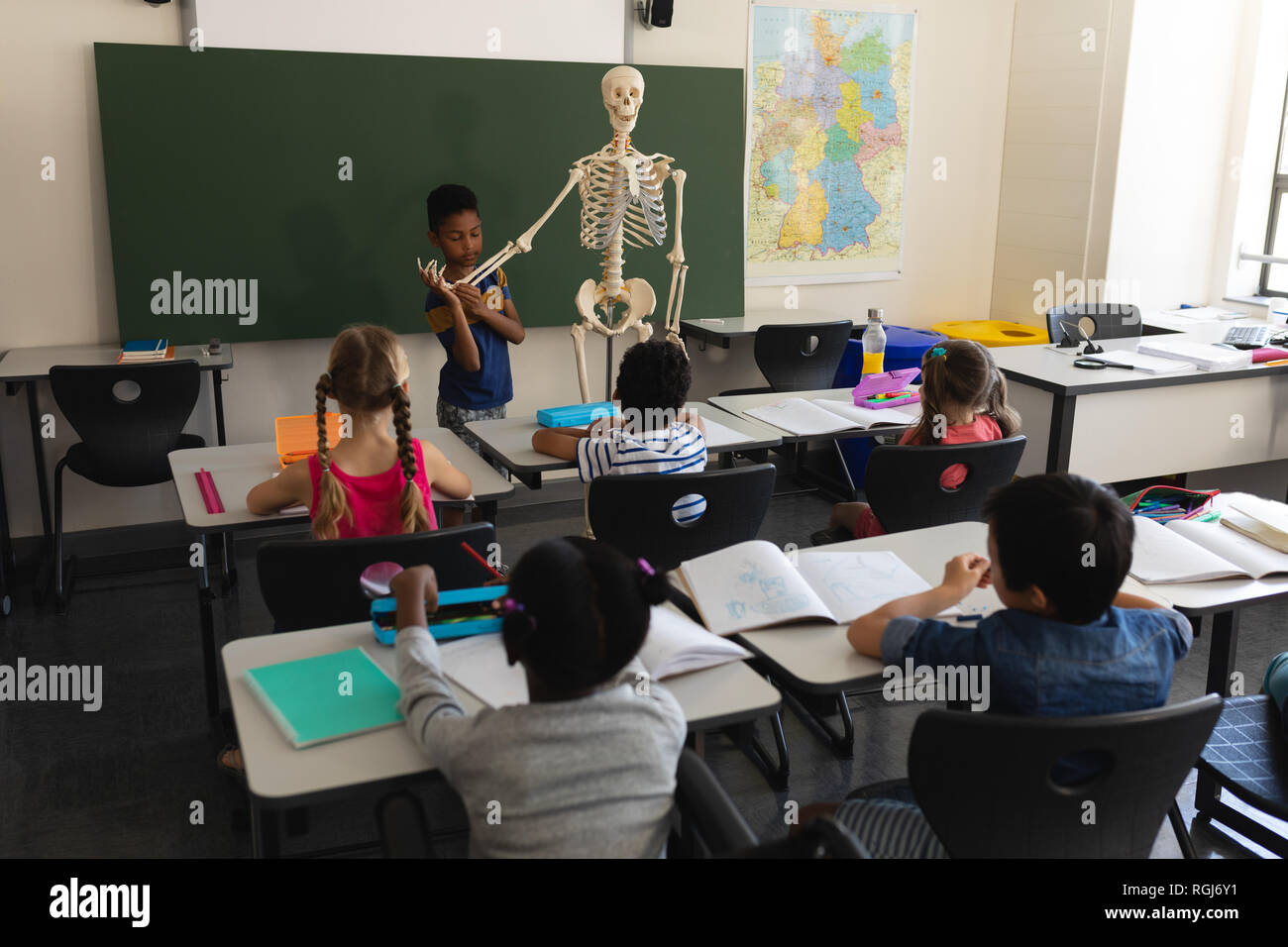 Rear view of schoolboy explaining human skeleton model in classroom of ...