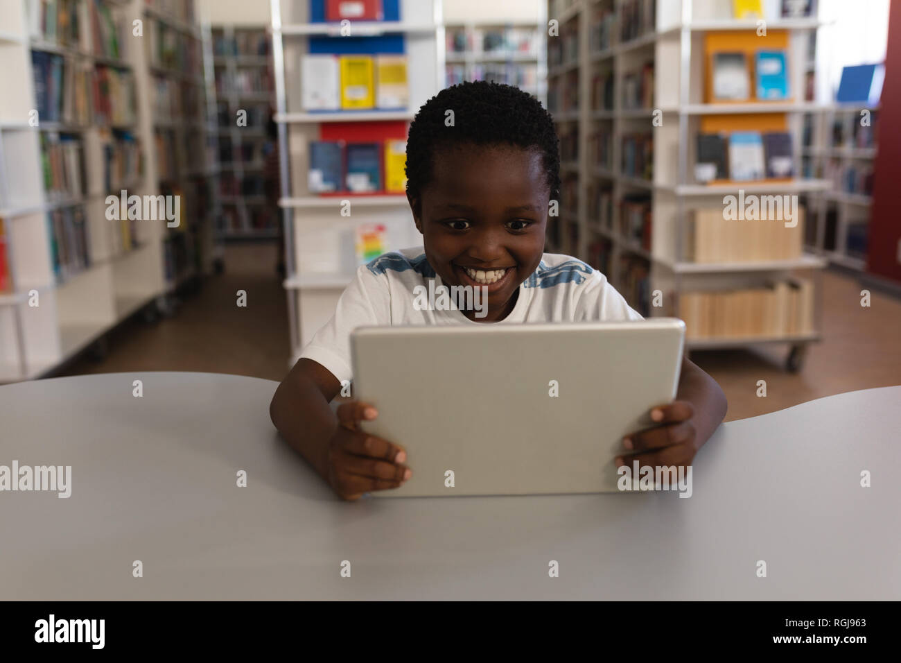 Happy schoolboy studying on digital tablet at table in school library ...