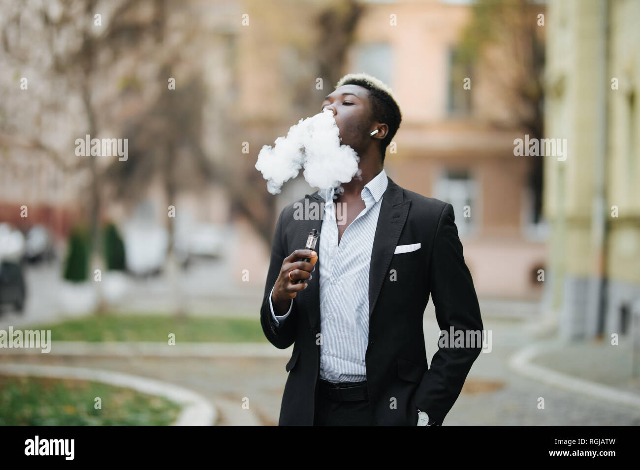 African man in a suit strolling on the streets of the city with vaping ...